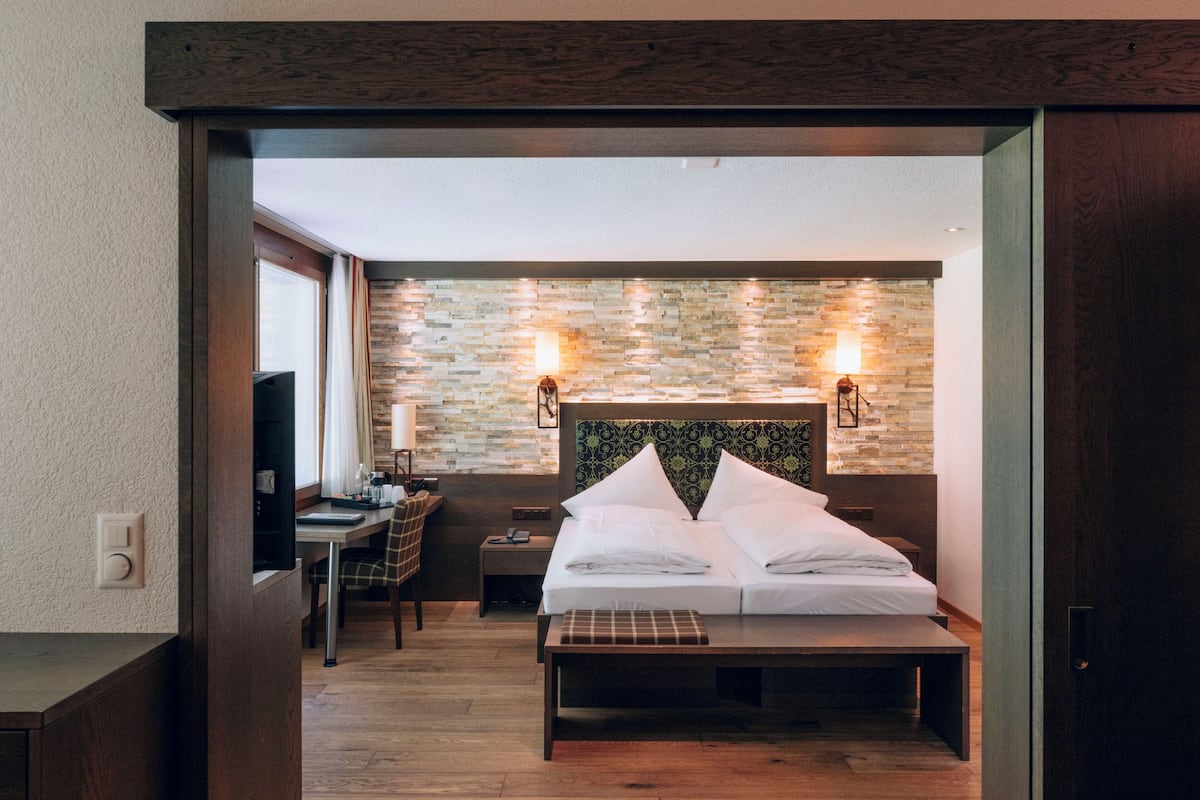 Quarto de hotel moderno com piso de madeira, cama com roupa branca, um banco, uma escrivaninha com cadeira e parede de pedra com duas luminárias de parede.