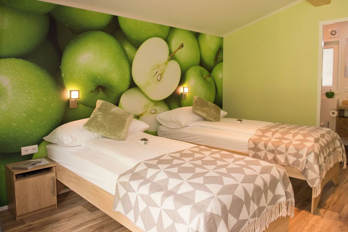Quarto de hotel com duas camas, um mural de uma maçã verde na parede e uma pequena mesa de cabeceira de madeira.