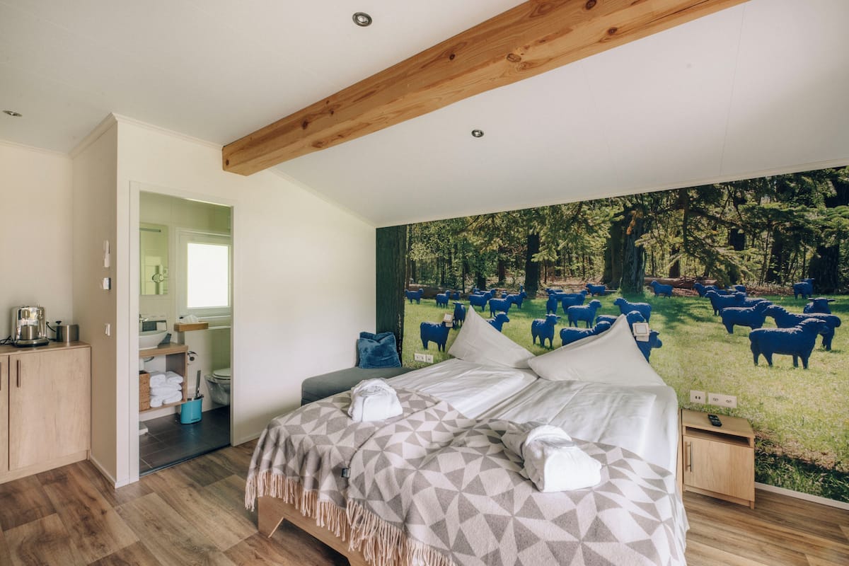 Quarto com uma cama grande, teto de madeira e um mural de um campo com ovelhas.
