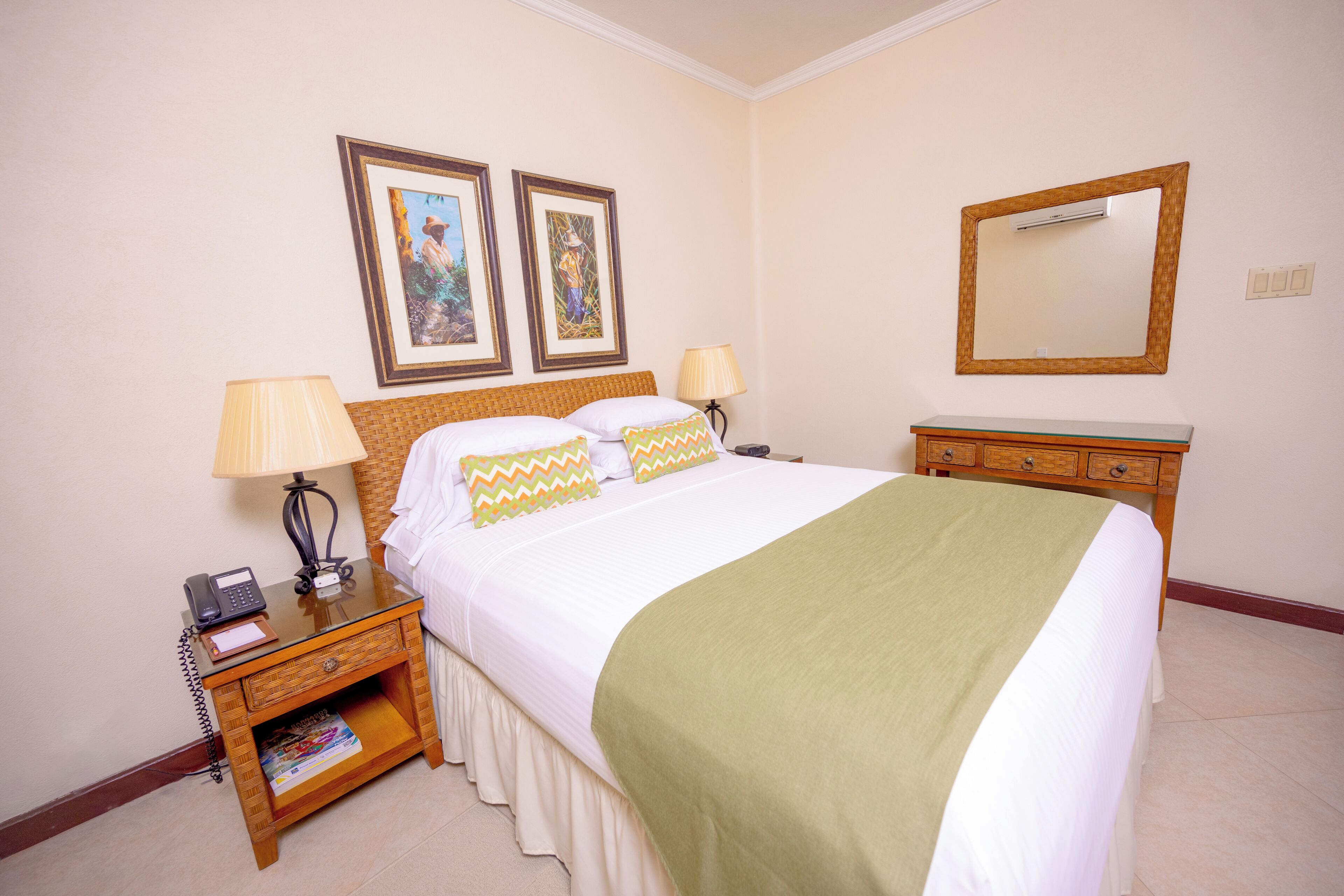Quarto de hotel bem arrumado com uma cama, abajures, mesa de cabeceira com telefone, uma revista e duas fotos emolduradas na parede.
