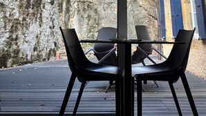 Uma área de refeições com mesa e cadeiras, vista para um penhasco rochoso e um edifício com persianas azuis.