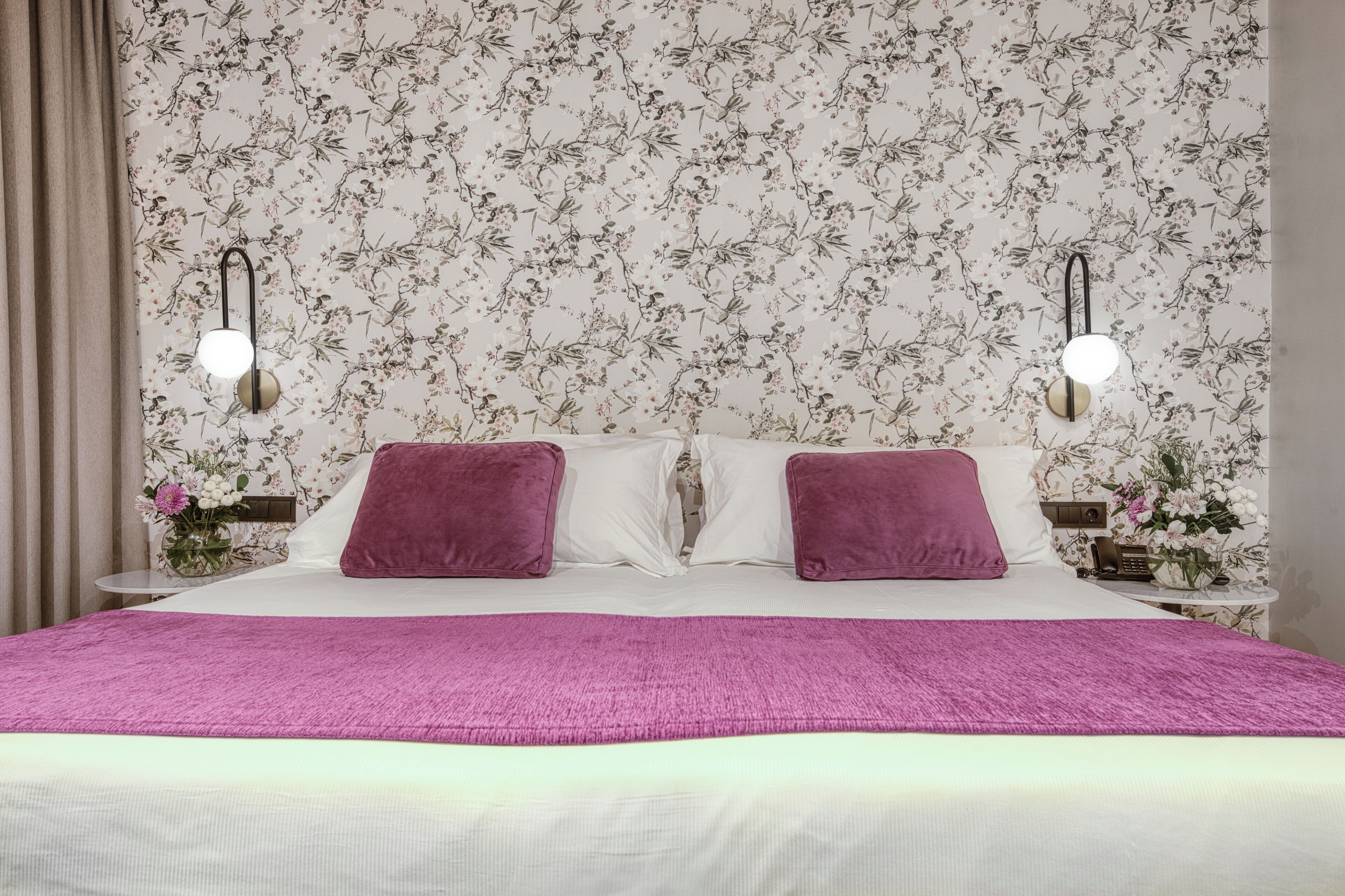 Quarto de hotel com papel de parede floral, duas almofadas roxas, um cobertor rosa e mesas de cabeceira com abajures.