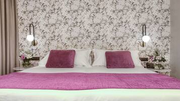 Quarto de hotel com papel de parede floral, duas almofadas roxas, um cobertor rosa e mesas de cabeceira com abajures.