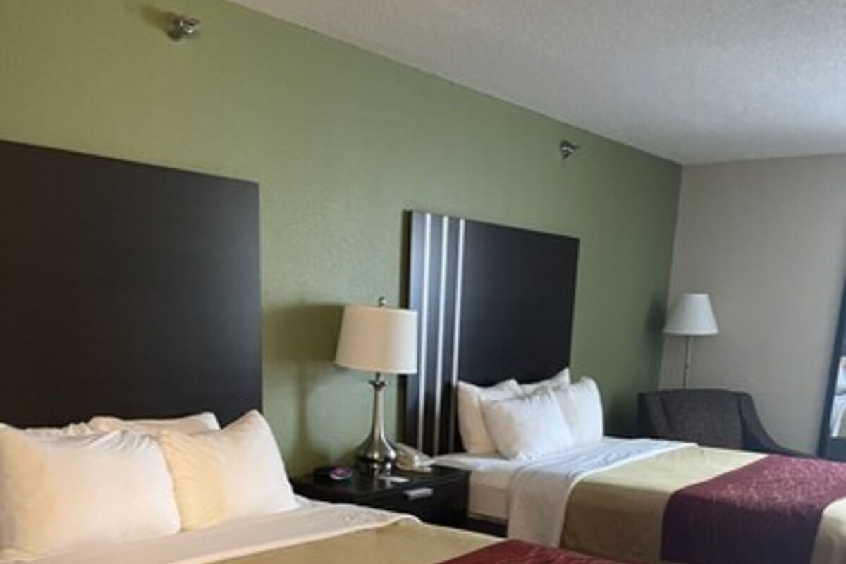 Quarto de hotel com duas camas, mesa de cabeceira, abajur e uma cadeira.