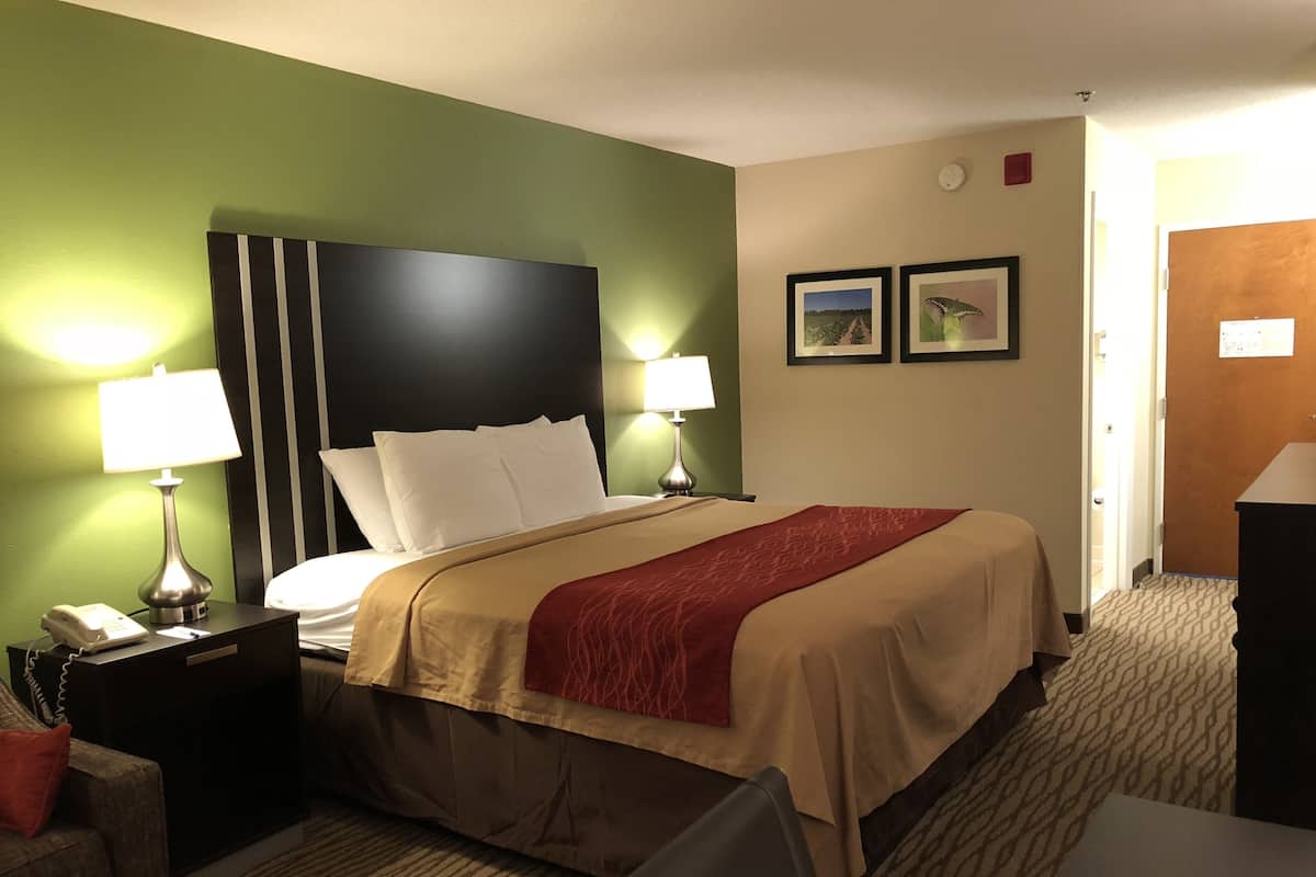 Quarto de hotel com uma cama, duas luminárias de cabeceira, um telefone, uma escrivaninha, uma cadeira e uma porta.