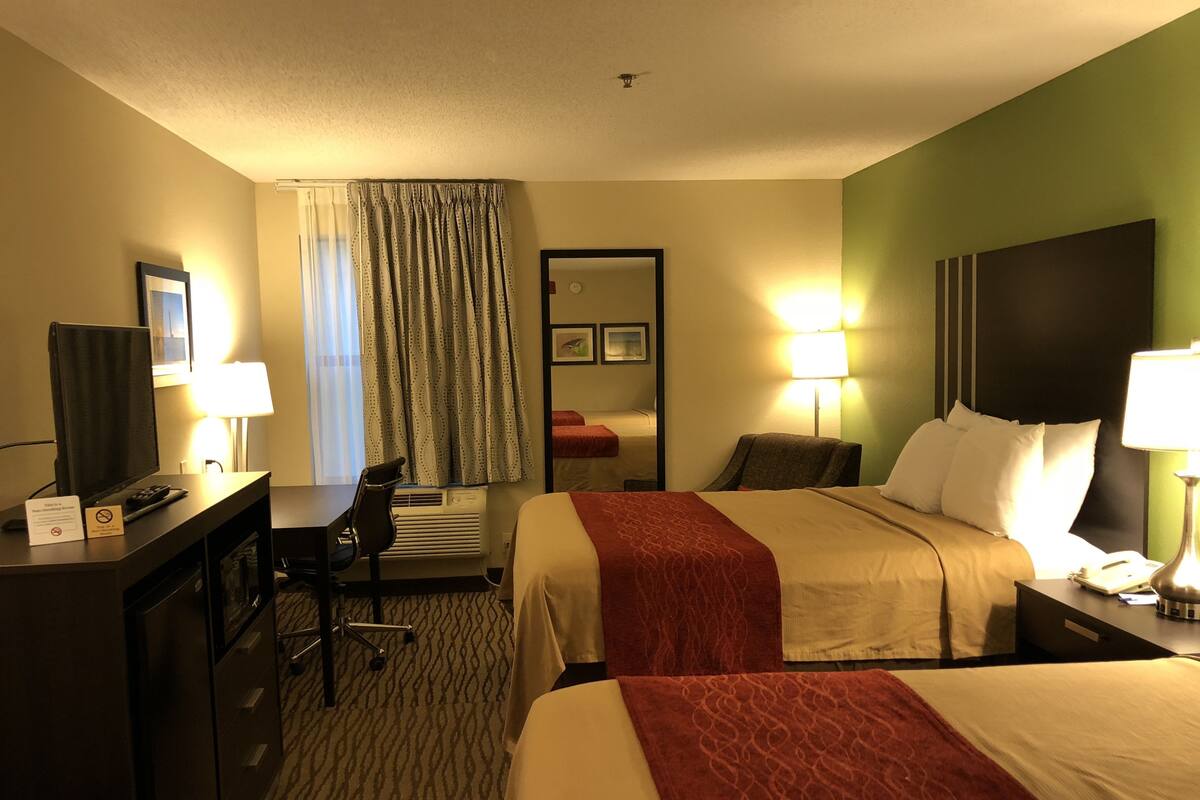 Quarto de hotel com duas camas, uma escrivaninha com computador, uma cadeira e uma televisão.