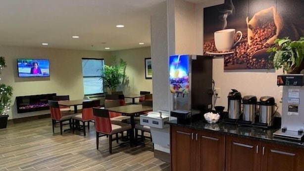 Quarto de hotel moderno com área de refeições, espaço para café e TV exibindo um telejornal.