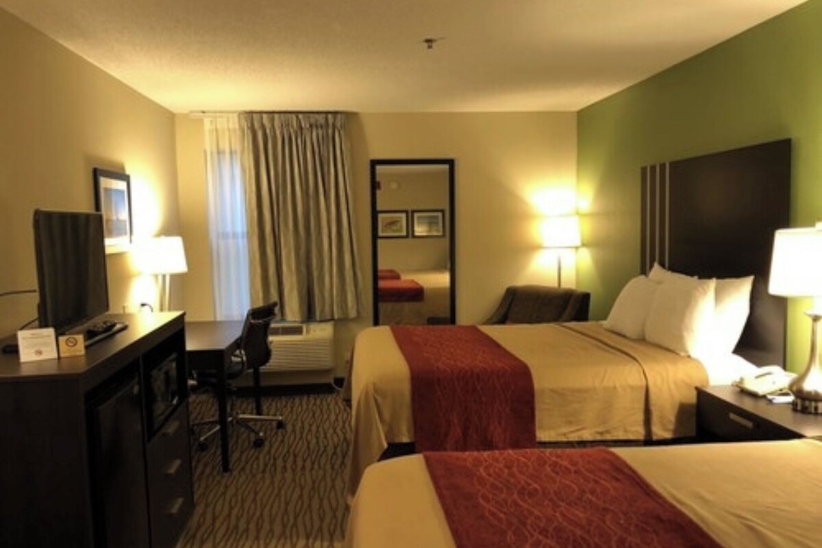 Quarto de hotel com duas camas, uma escrivaninha, uma cadeira, abajur de escrivaninha, luminária de cabeceira e um espelho grande.