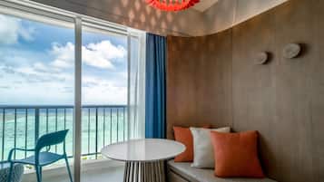 Uma varanda com vista para o oceano, uma mesa redonda, uma cadeira azul e um banco estofado com almofadas laranja.