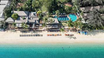 Vista aérea de um resort com uma grande piscina, espreguiçadeiras na praia e palmeiras.