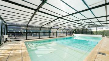 Piscina interior com teto de vidro, chão azulejado e vista para o exterior.