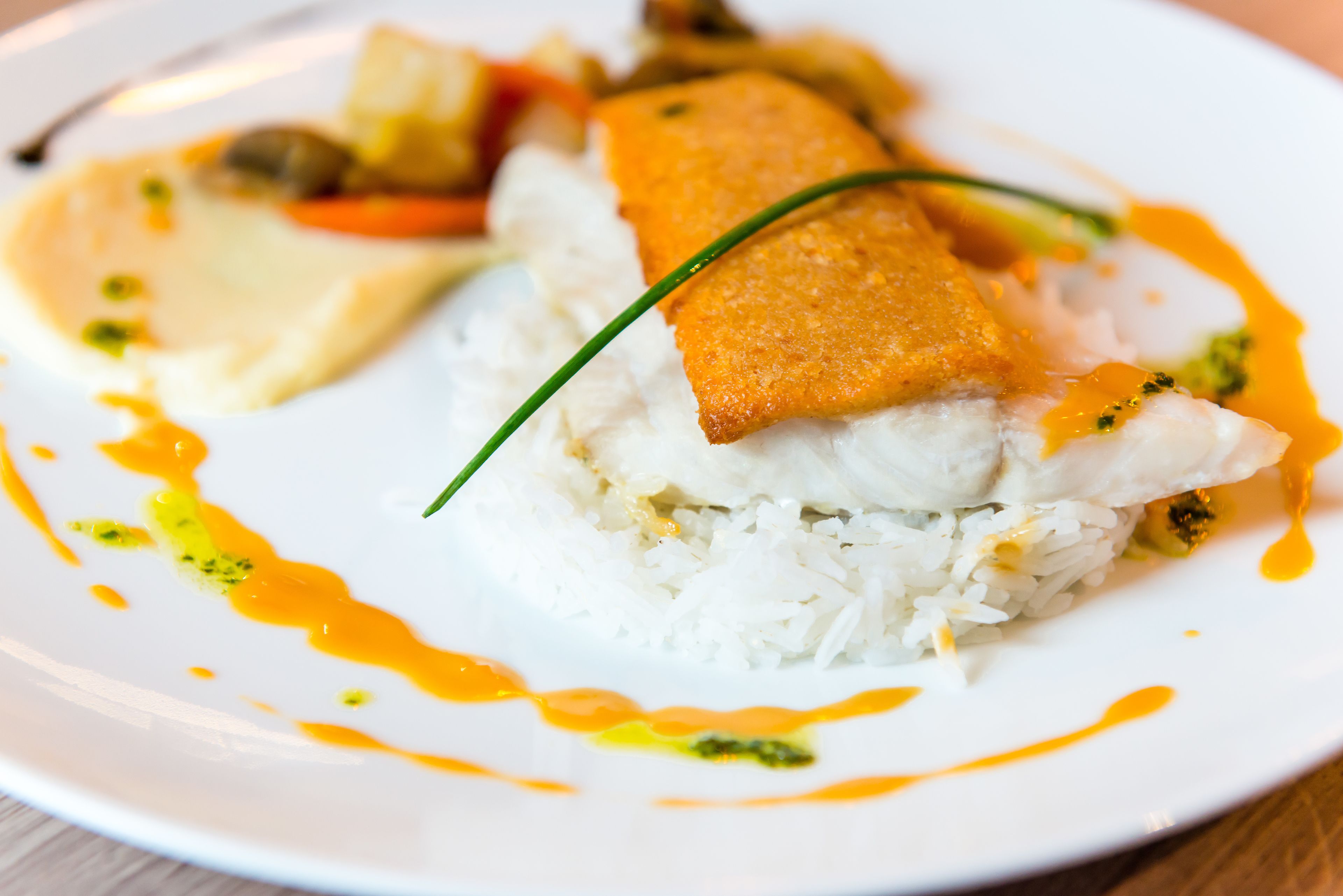Prato de arroz branco coberto com um pedaço de peixe e uma cobertura crocante, acompanhado de legumes e um fio de molho.