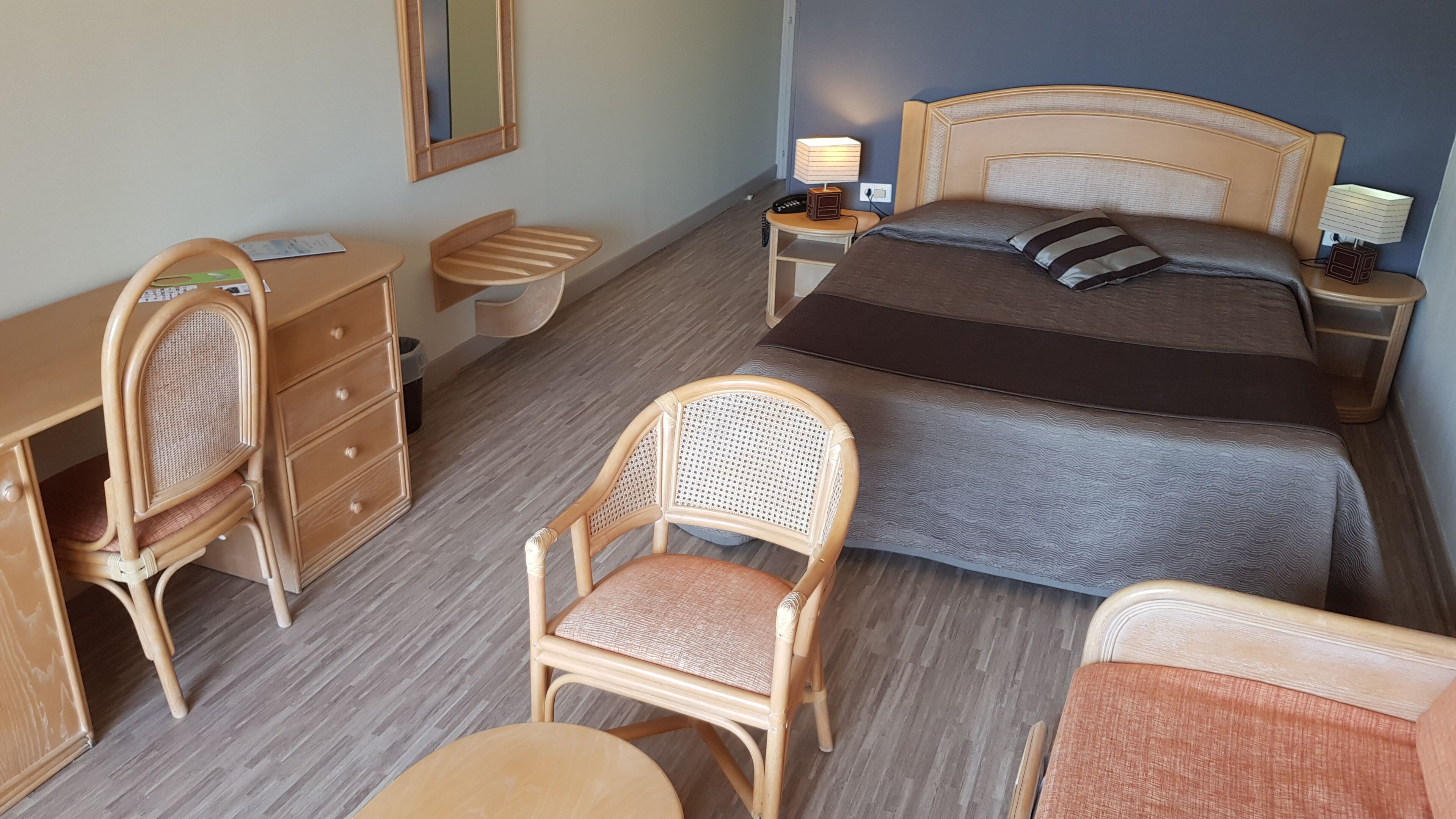 Quarto de hotel com cama, duas cadeiras, escrivaninha, mesa de cabeceira com abajur e espelho.