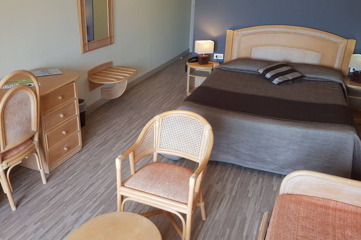Quarto de hotel com cama, duas cadeiras, escrivaninha, mesa de cabeceira com abajur e espelho.