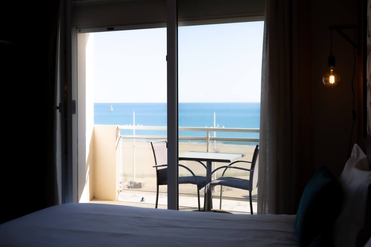 Quarto de hotel com cama, mesa e cadeiras perto de uma porta de correr de vidro que dá para uma varanda com vista para o mar.
