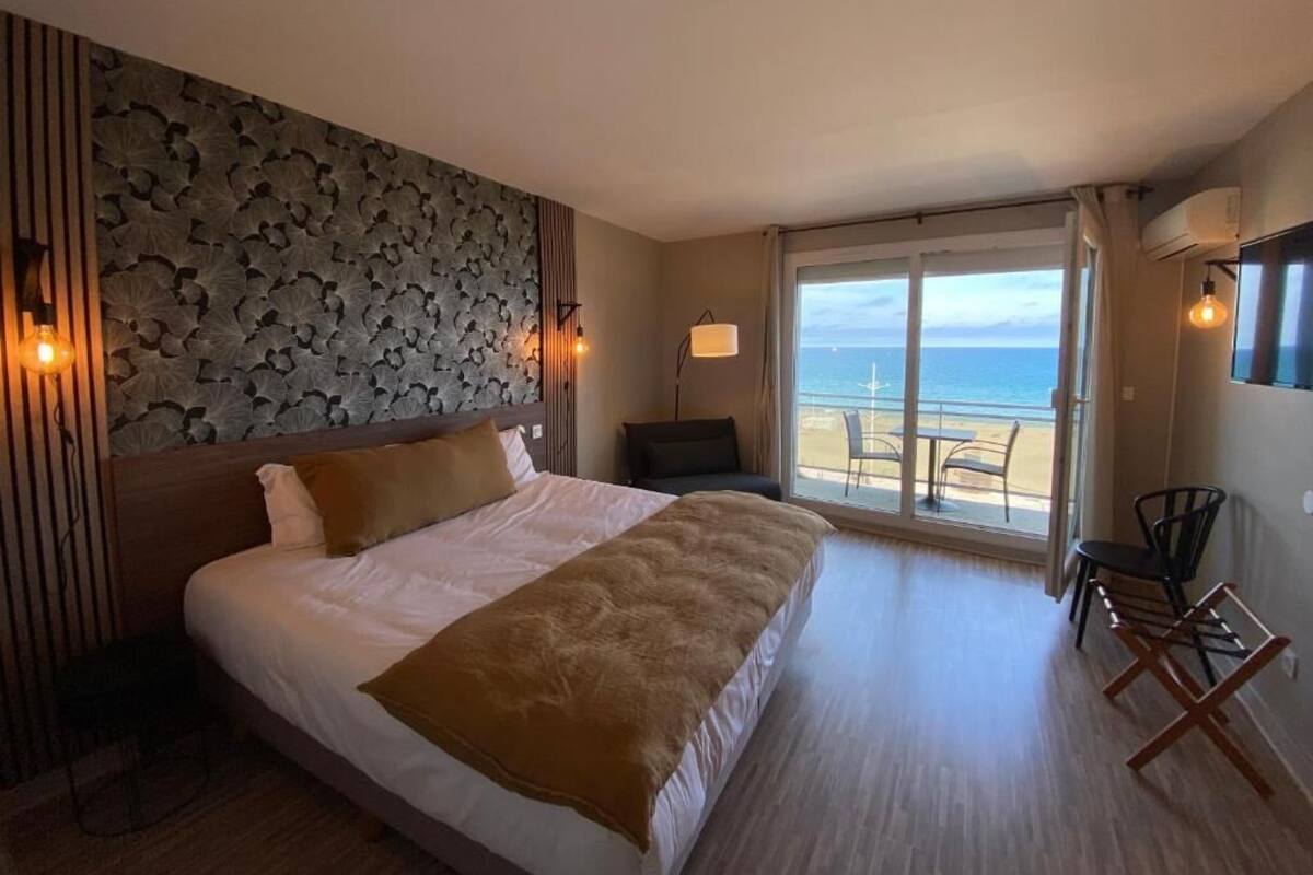 Quarto de hotel com uma cama grande, vista para a praia, escrivaninha com cadeira e televisão.