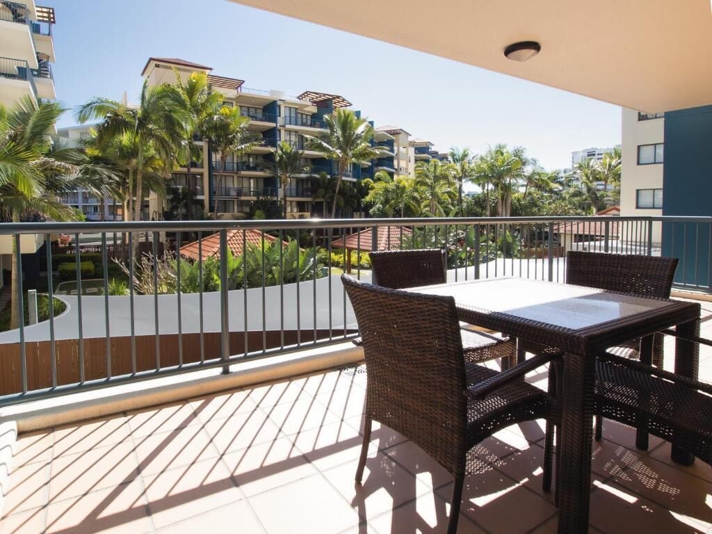 Uma varanda com mesa e cadeiras, com vista para uma área de piscina, palmeiras e edifícios ao fundo.