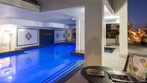 Uma piscina interior com piso e paredes revestidas, uma mesa de madeira com revistas, e vista para a cidade à noite.