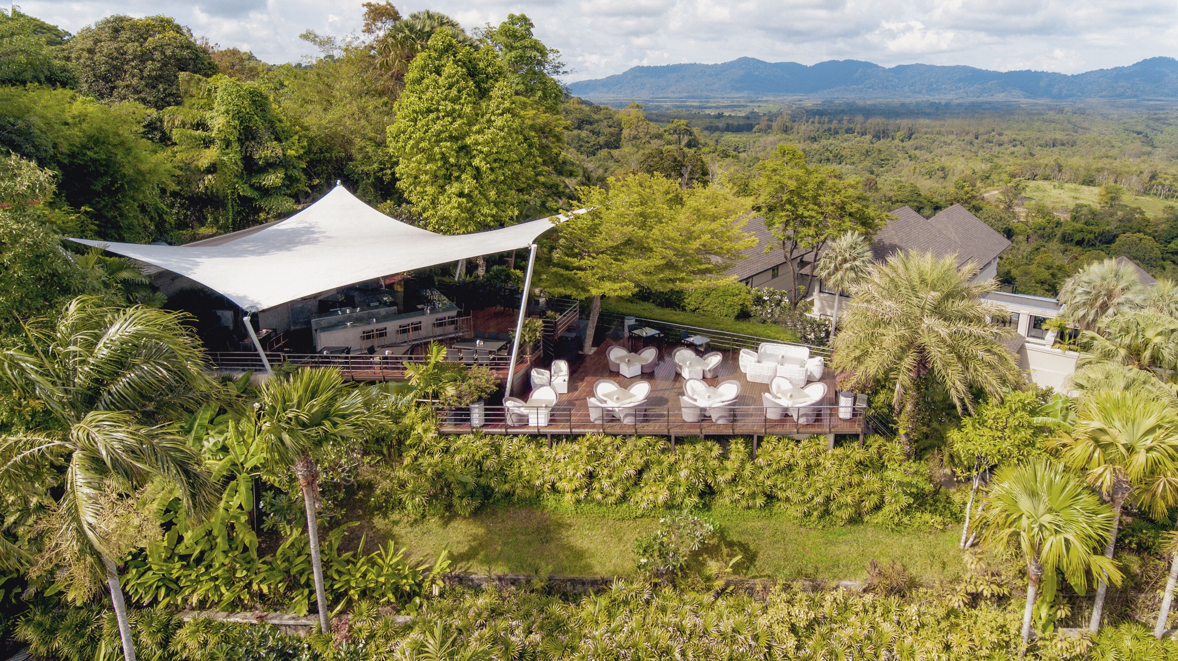 Um resort com uma ampla área externa coberta, com mobiliário branco, cercado por vegetação exuberante e palmeiras.