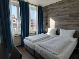 Quarto de hotel com duas camas, cabeceira de madeira, vista para edifícios e janela com cortinas azuis.