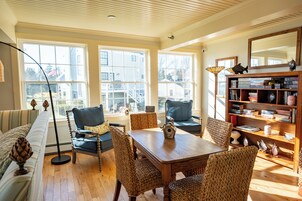 Um quarto acolhedor com uma mesa de jantar de madeira, cadeiras de vime, uma estante de livros e grandes janelas com vista para edifícios vizinhos.