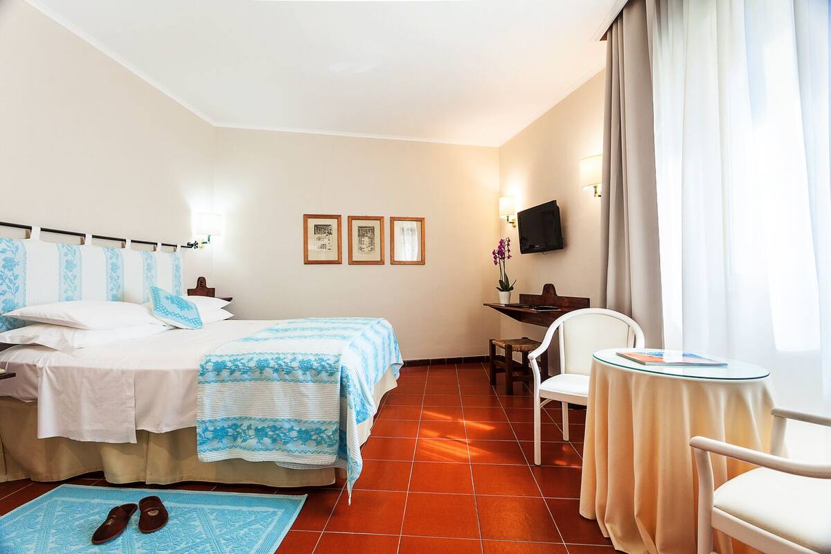 Quarto de hotel com cama, escrivaninha, cadeira, televisão e janela com cortinas.
