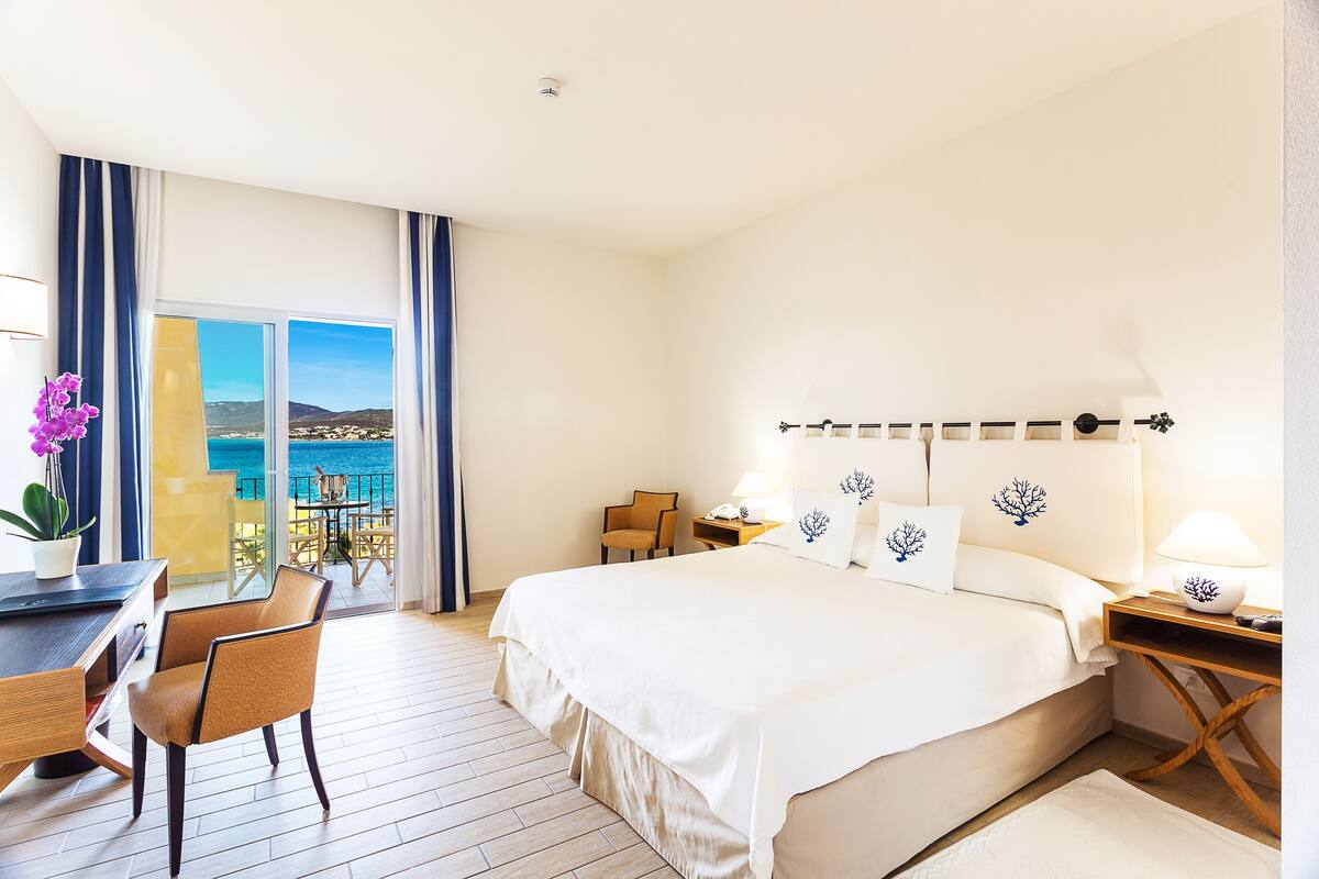 Quarto de hotel com uma cama grande, uma mesa, uma cadeira, uma mesa de cabeceira e vista para o mar.