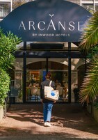 Uma pessoa caminhando em direção à entrada de um hotel chamado 'ARCÂNSE BY INWOOD HOTELS'.