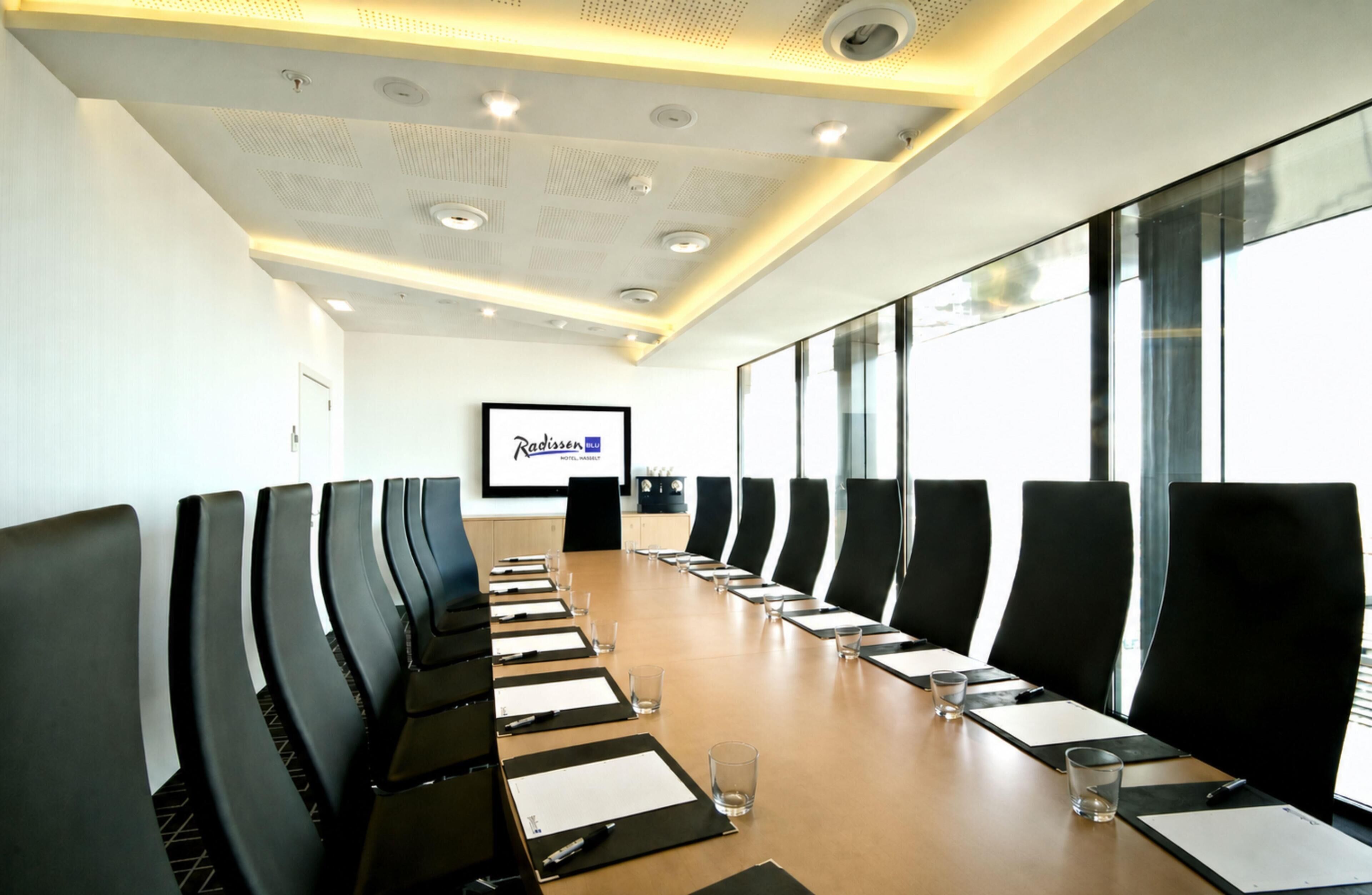 Uma sala de conferências com uma mesa comprida, cadeiras pretas e uma tela exibindo 'Radissen BLDG'.