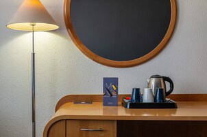 Quarto de hotel com um espelho redondo, uma lâmpada, uma cafeteira e um tabuleiro com xícaras e uma caixa.