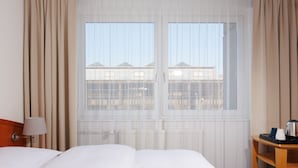 Quarto de hotel com uma cama grande, uma mesa de cabeceira com abajur e vista para edifícios através de cortinas leves.