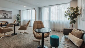 Quarto de hotel moderno com sofá, poltronas, mesa de vidro e uma janela grande com cortinas translúcidas.