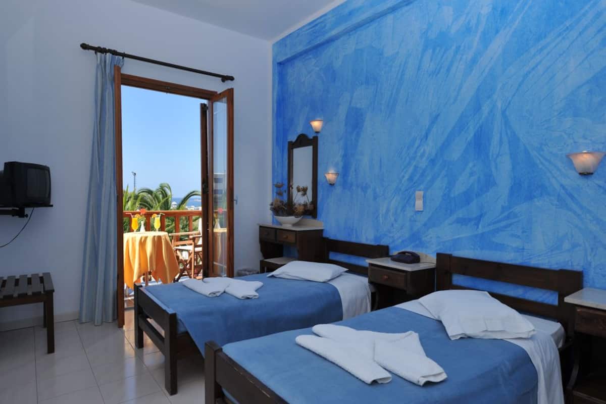 Quarto de hotel com duas camas, varanda com vista, televisão e parede azul grande.