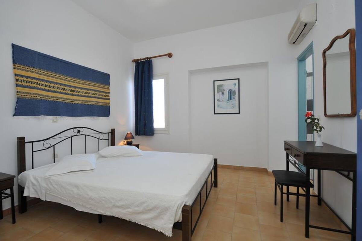 Quarto com cama, escrivaninha, cadeira, janela com cortinas, ar-condicionado fixado na parede e um quadro emoldurado.