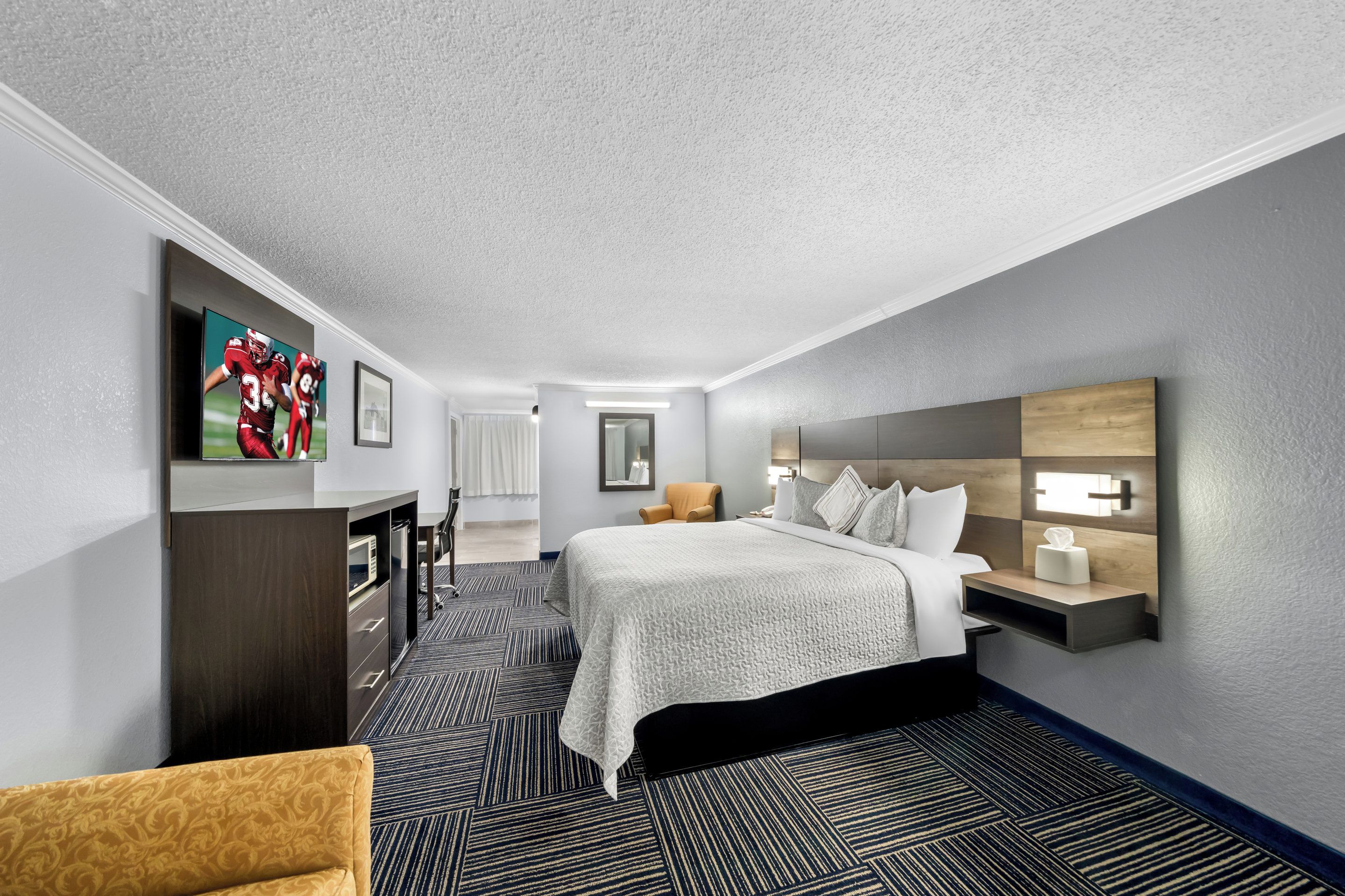 Quarto de hotel com uma cama grande, uma escrivaninha com televisão de ecrã plano, uma cadeira, uma mesa de cabeceira com abajur e uma estante na parede.