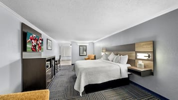 Quarto de hotel com uma cama grande, uma escrivaninha com televisão de ecrã plano, uma cadeira, uma mesa de cabeceira com abajur e uma estante na parede.