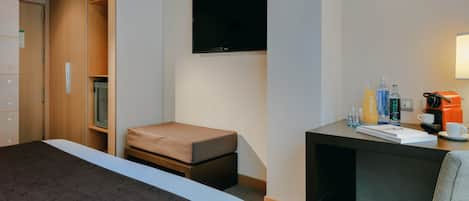Quarto de hotel com cama, pequena escrivaninha com cafeteira, televisão na parede e guarda-roupa.