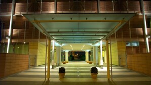 Entrada coberta a um edifício, com piso xadrez, duas plantas em vasos e um caminho central que leva a uma porta.
