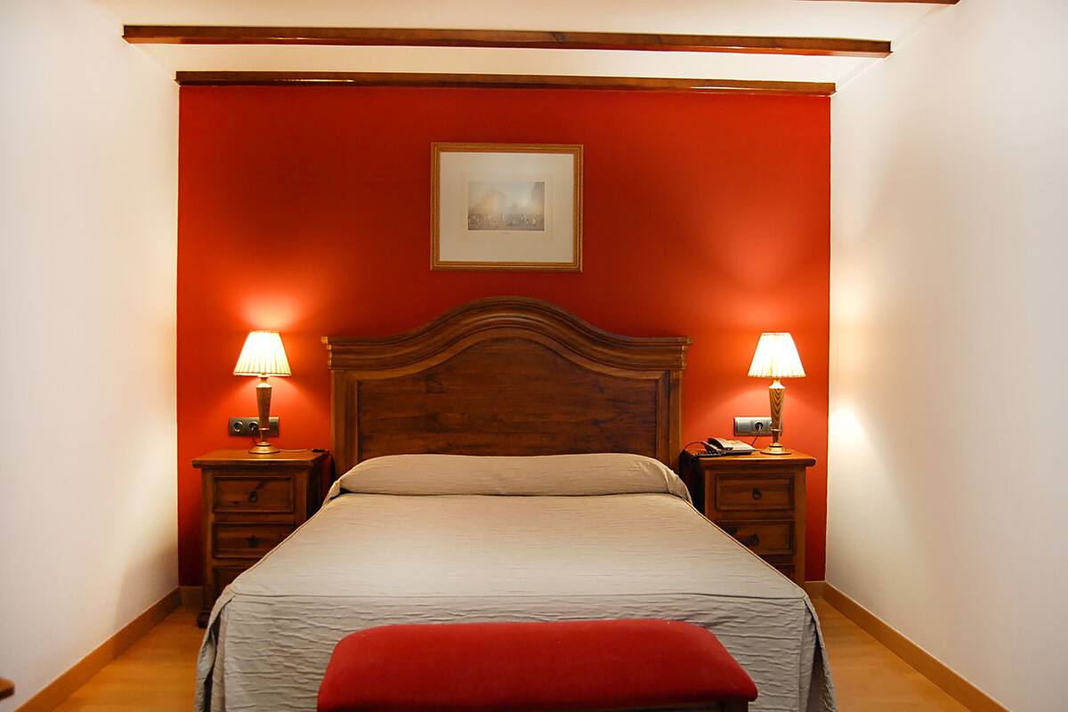 Quarto com cabeceira de madeira, duas mesas de cabeceira com luminárias, parede vermelha e um quadro emoldurado acima da cabeceira.