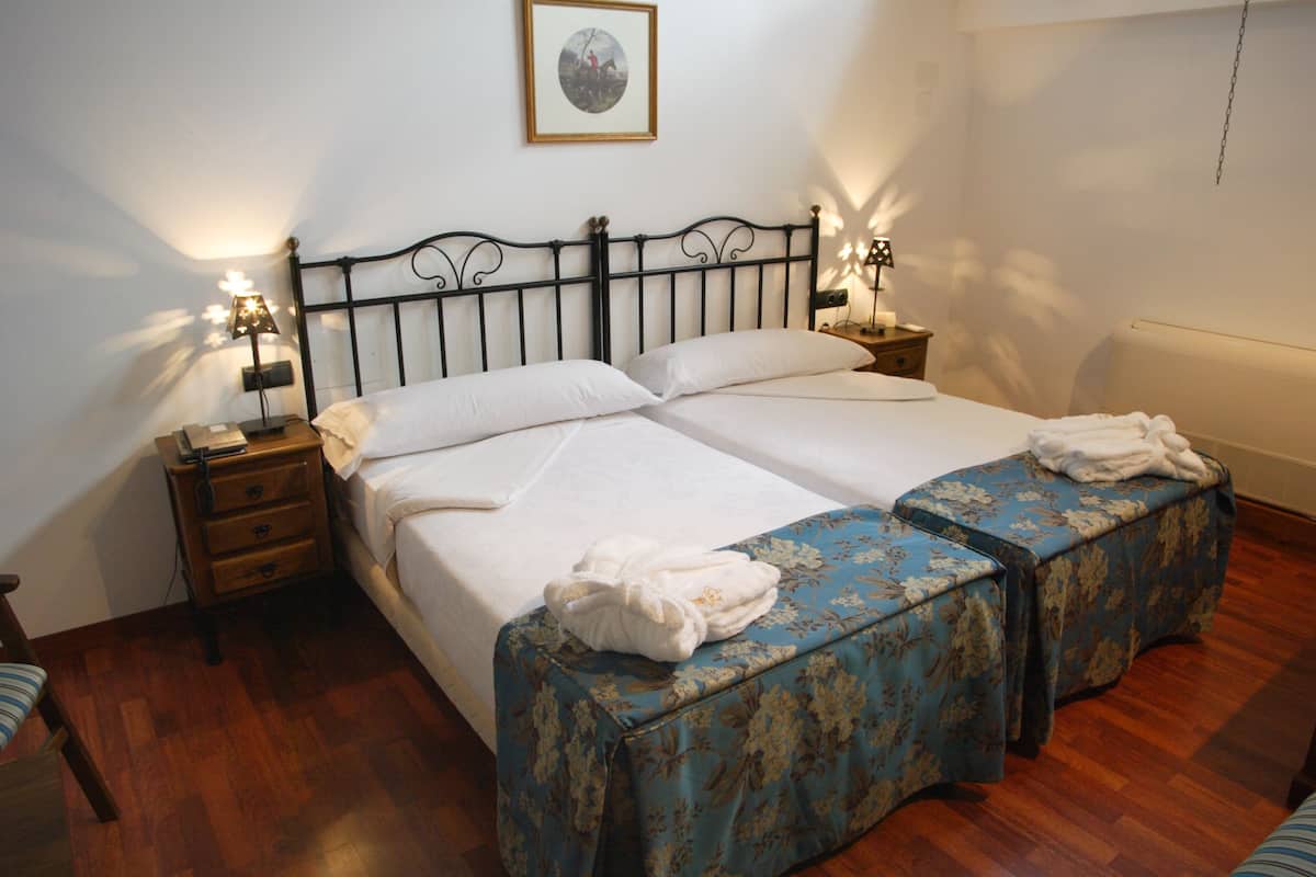 Cama de casal com roupa de cama branca, duas mesas de cabeceira com luminárias e um banquinho com padrão floral.