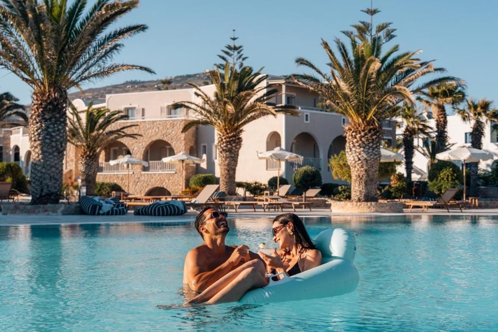 Um casal em uma piscina, com espreguiçadeira e guarda-sol, cercado por palmeiras e um edifício com arcos.