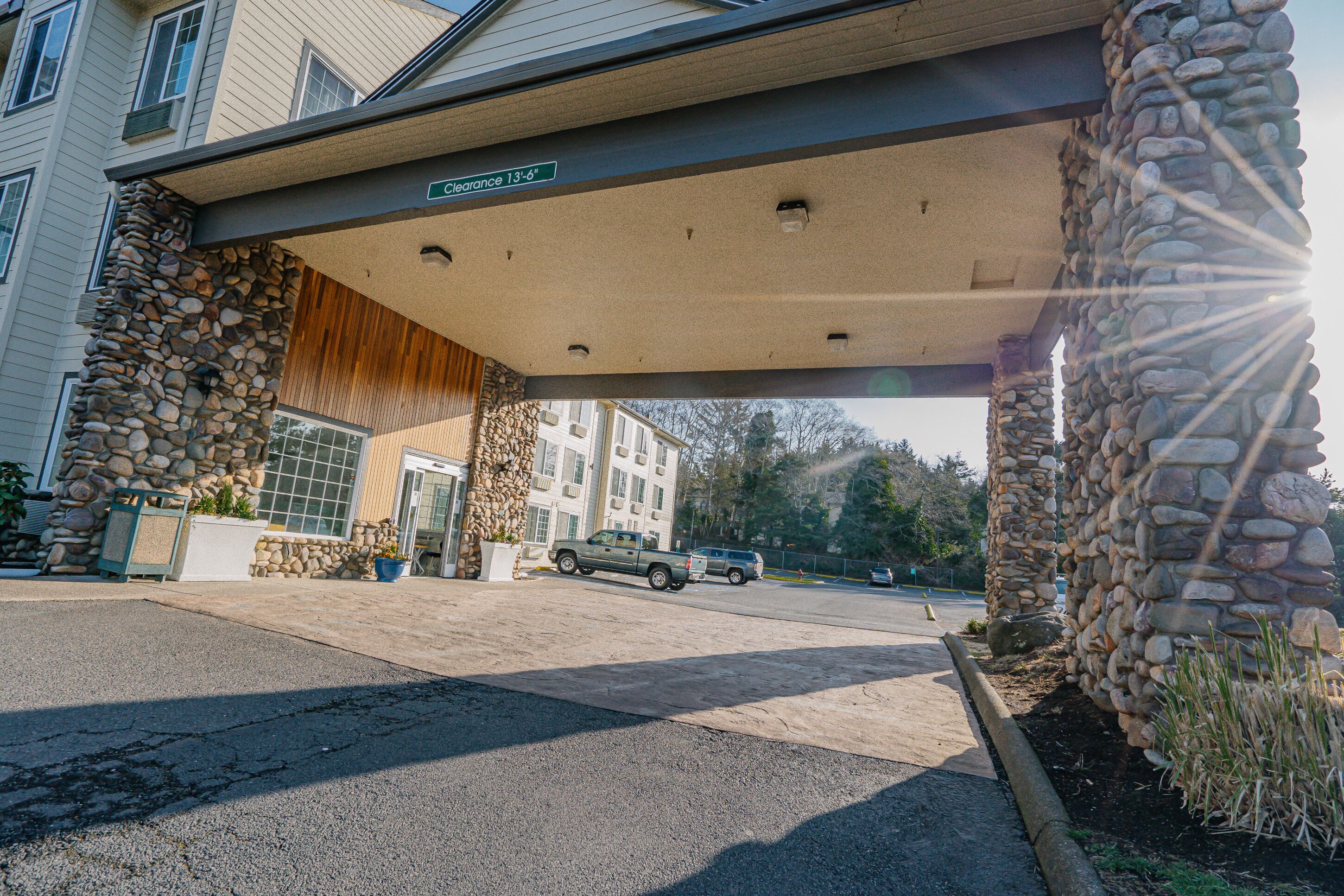 Entrada de hotel com pilares de pedra, galeria coberta e área de estacionamento.