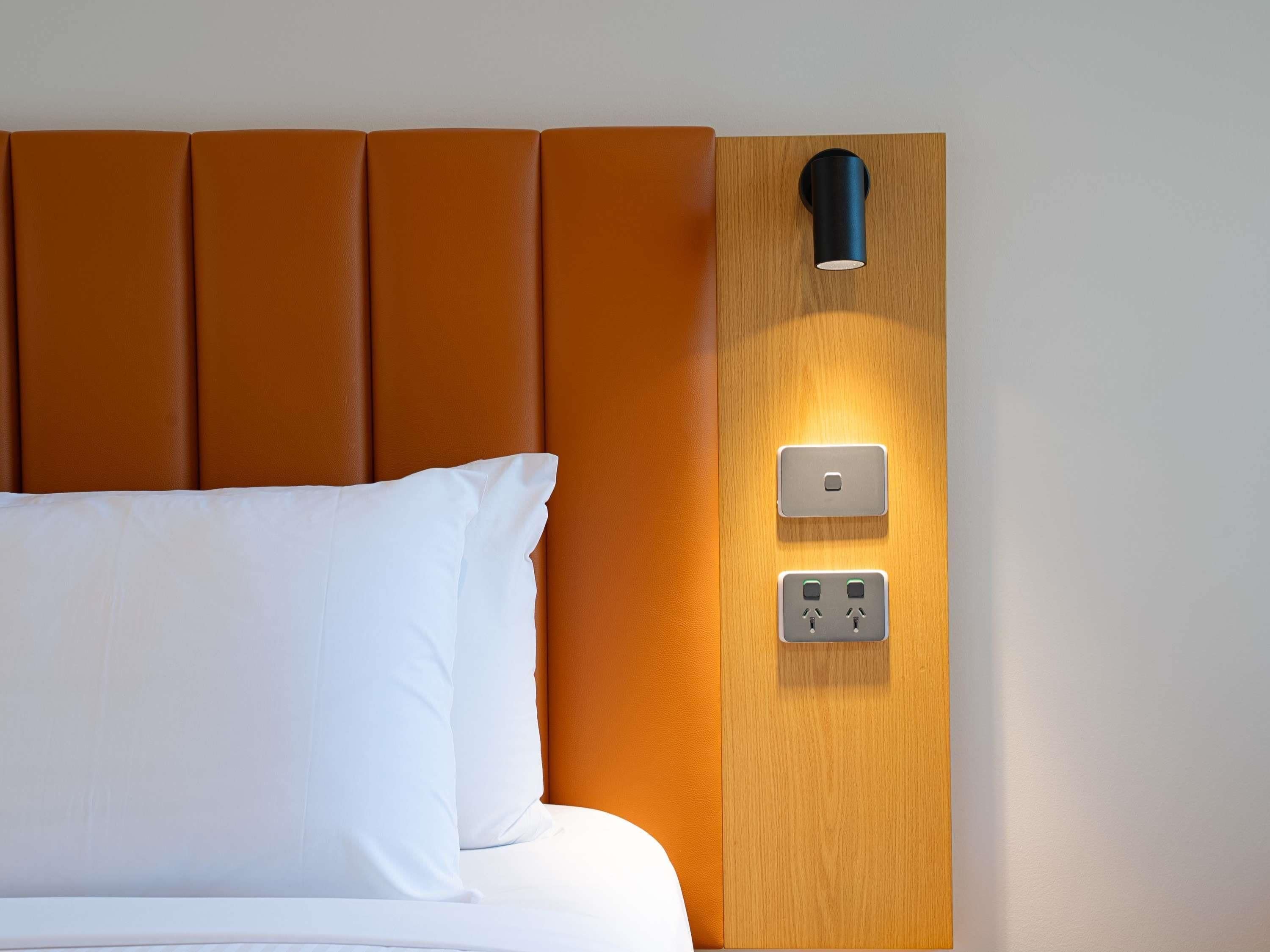 Cama com roupa de cama branca e cabeceira com painel de madeira e luminária.