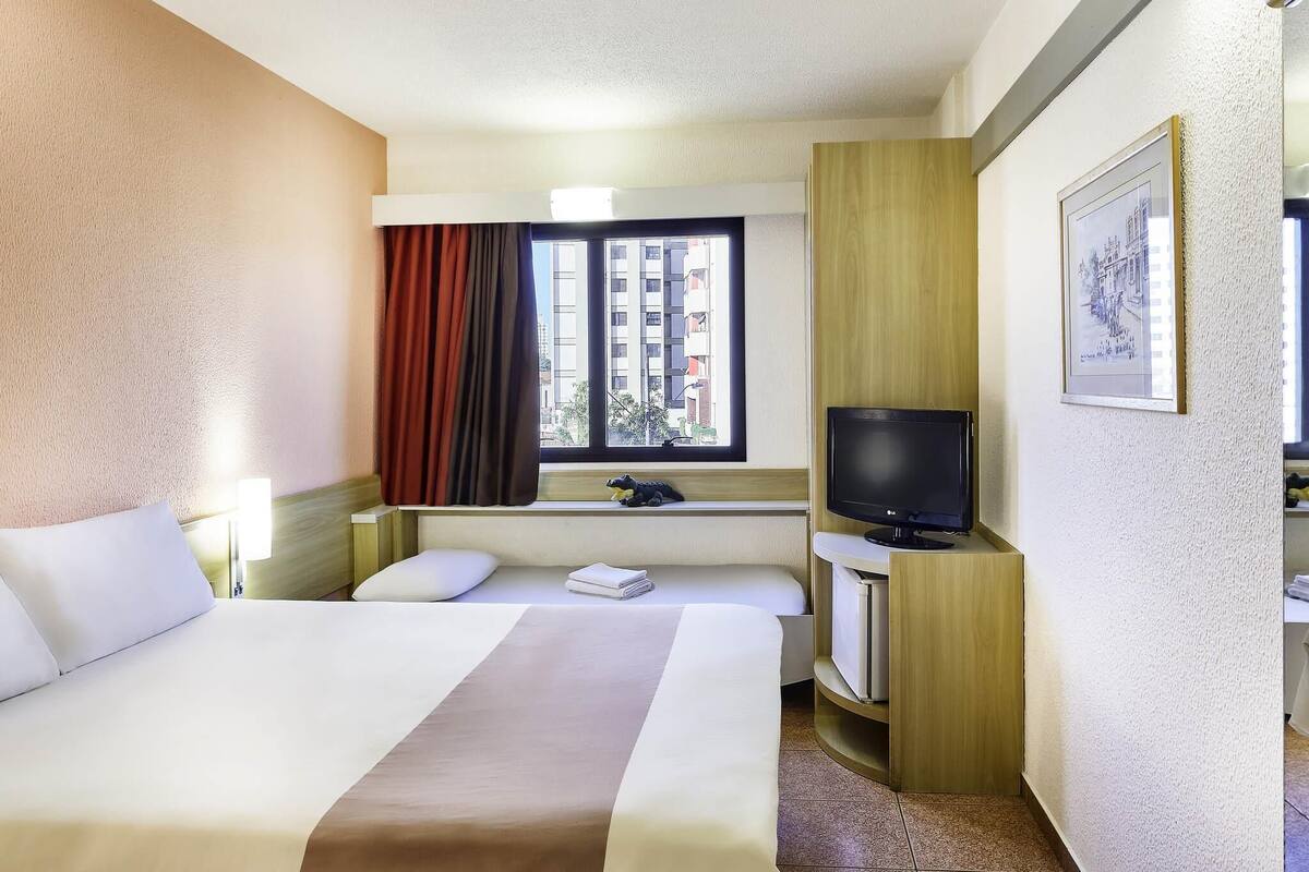 Quarto de hotel com cama, escrivaninha com computador, televisão e janela com cortinas.