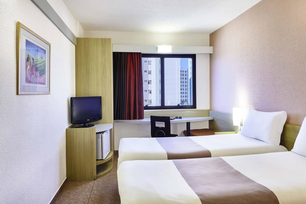Quarto de hotel com duas camas, uma escrivaninha, televisão e janela com cortinas.