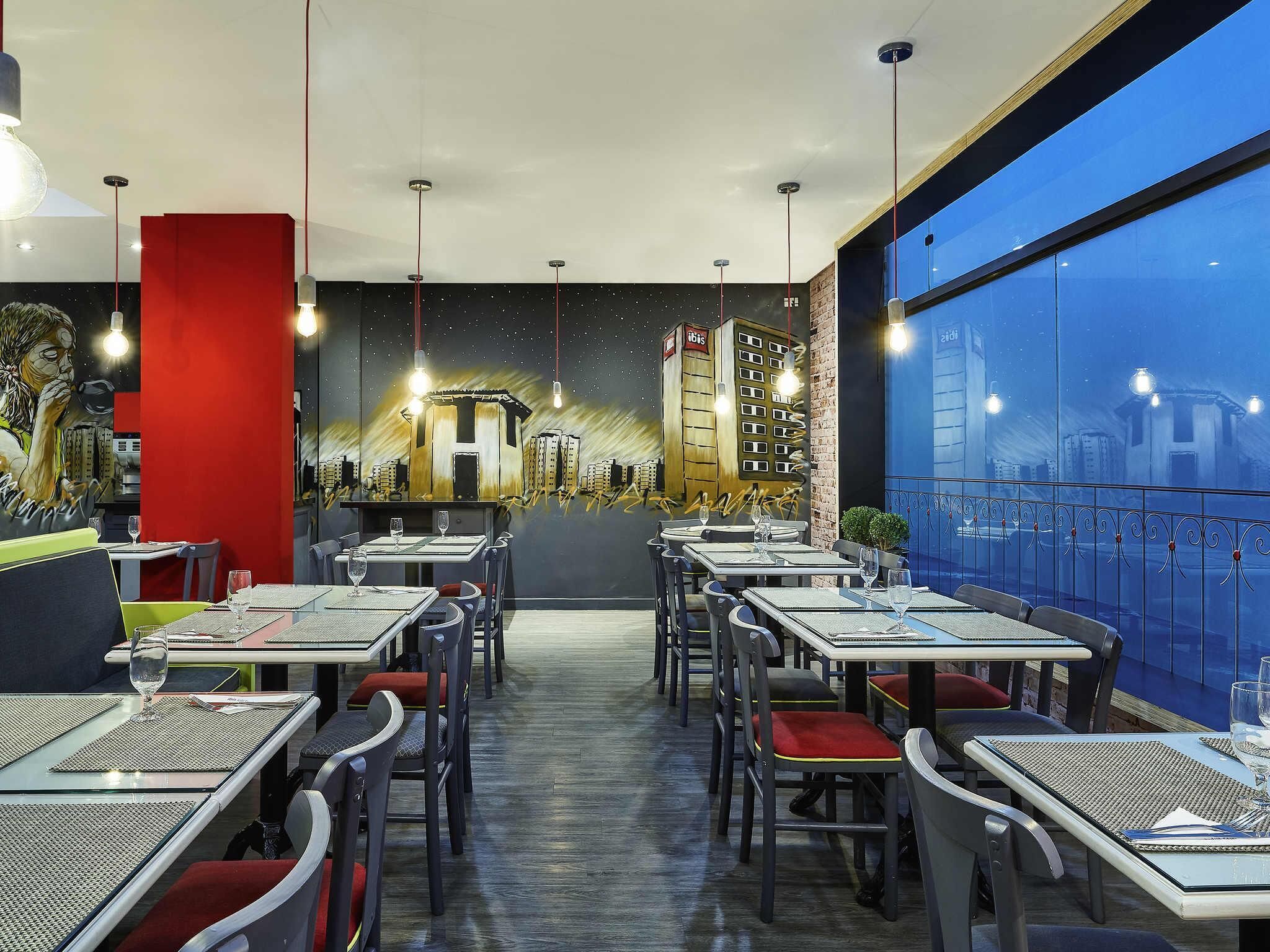 Um restaurante com mesas e cadeiras, um mural na parede e vista para prédios através de uma grande janela.