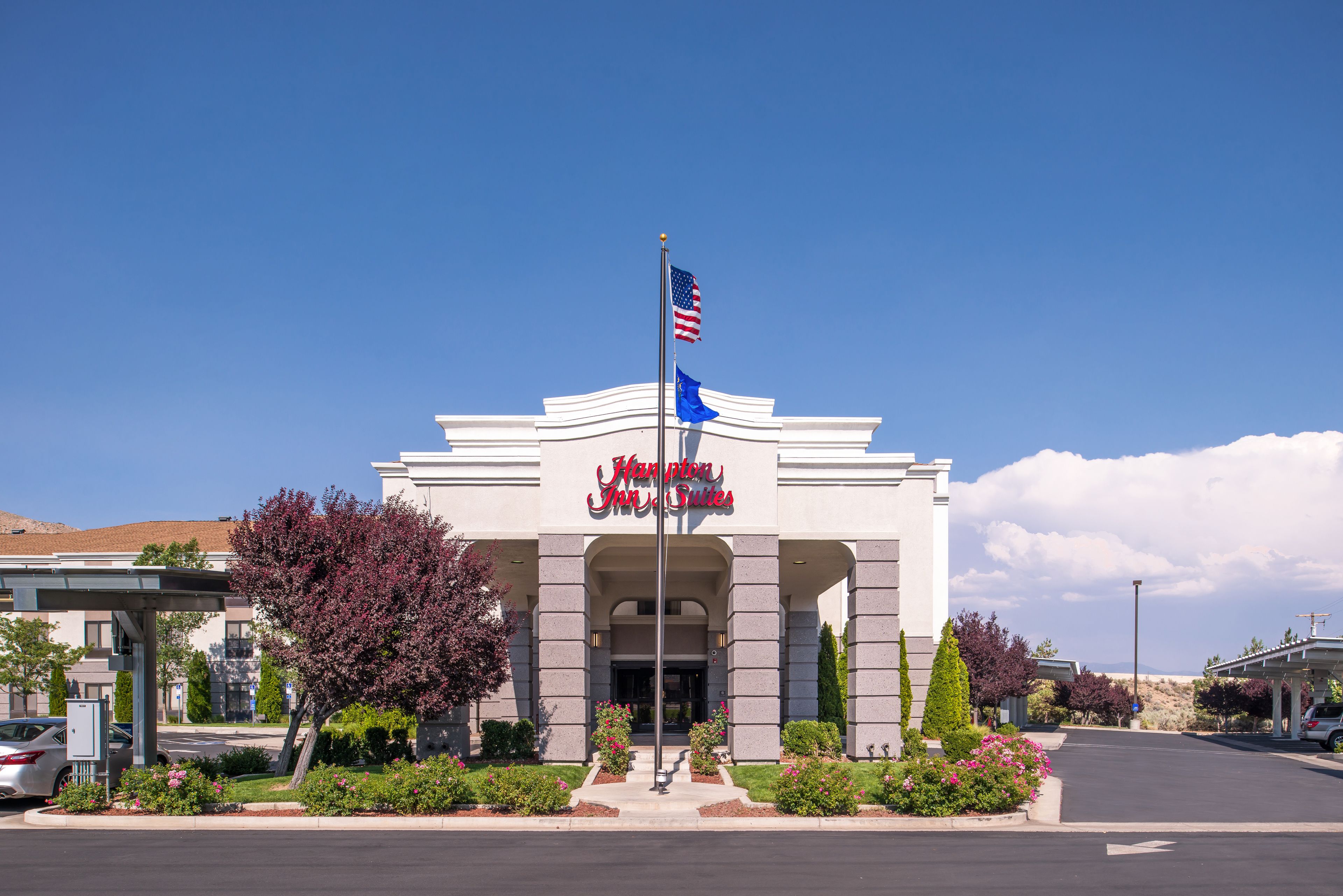 Entrada de hotel com céu claro, bandeira americana e uma placa com a inscrição 'Hampton Inn Suites'.