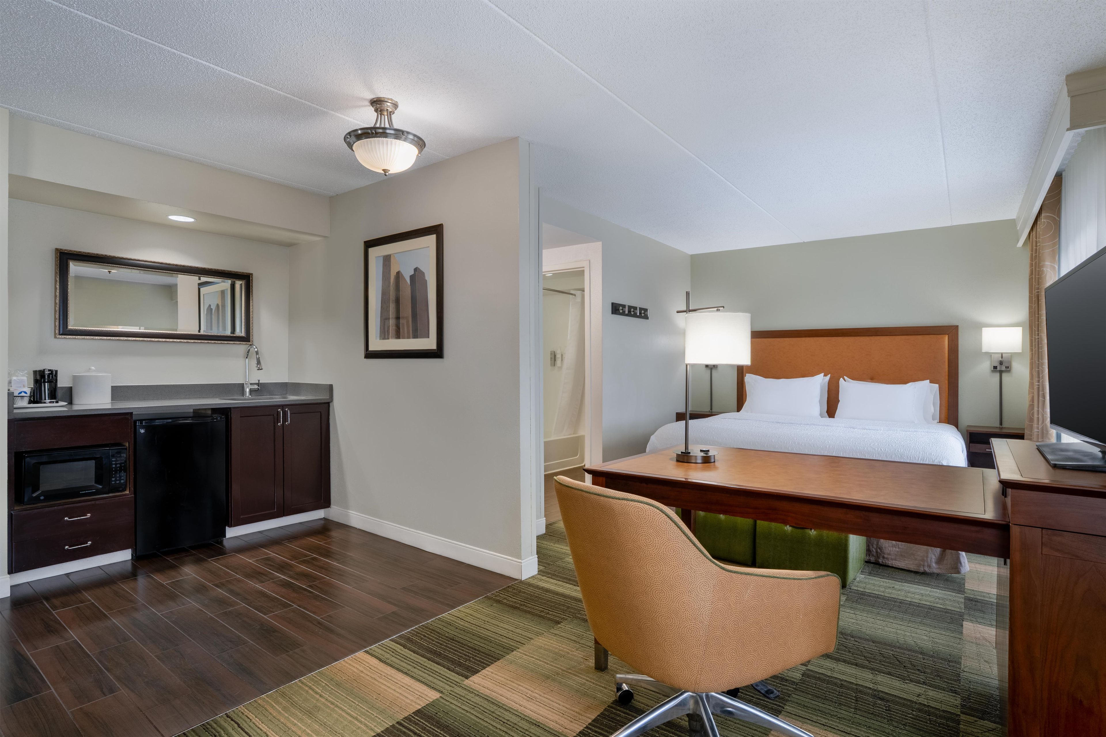 Quarto de hotel com cama, escrivaninha, cadeira e cozinha compacta.