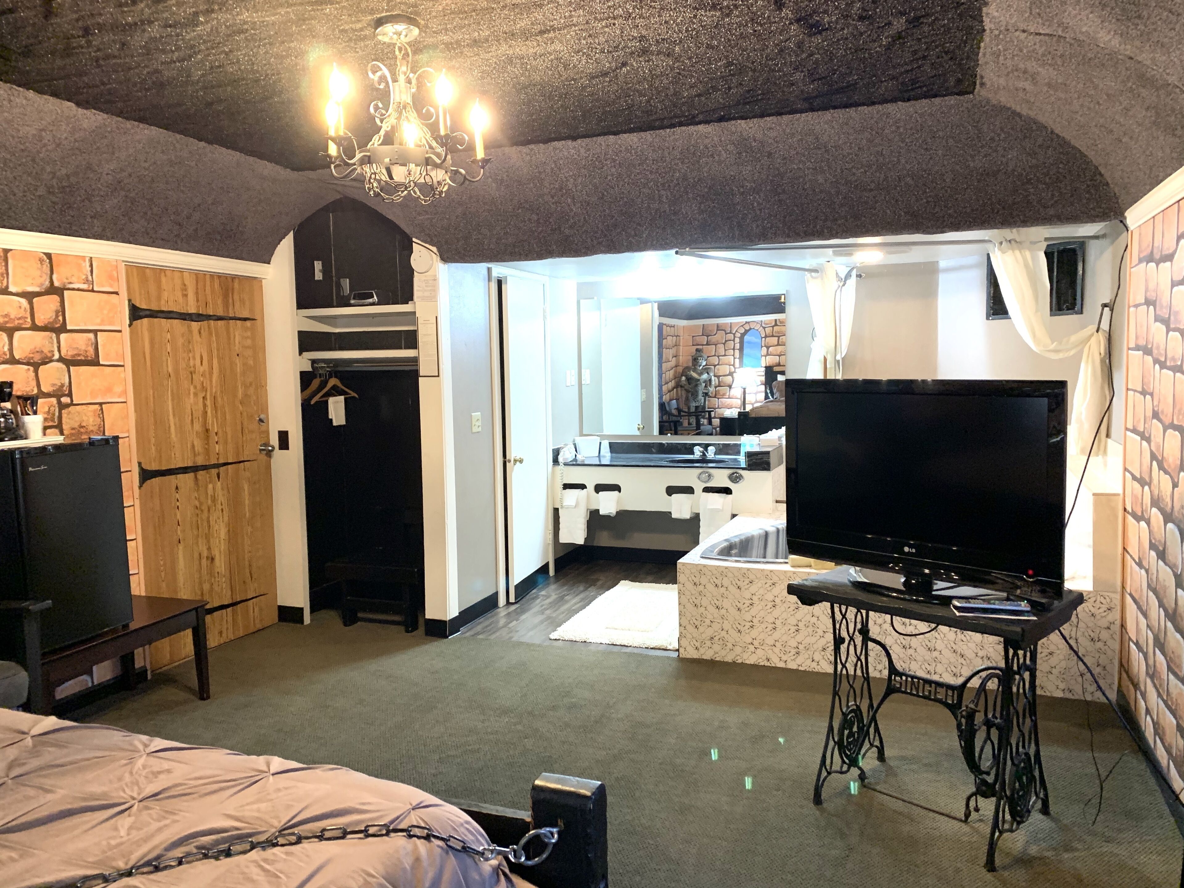 Quarto pequeno com cama, televisão, máquina de costura e cozinha compacta.