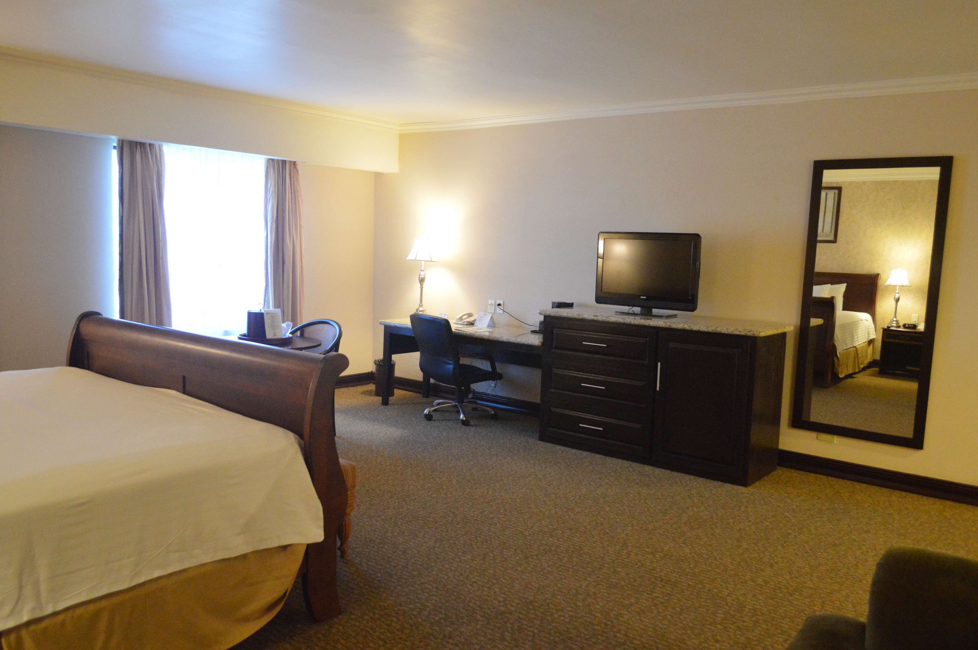Quarto de hotel com cama, escrivaninha, cadeira, televisão e espelho.